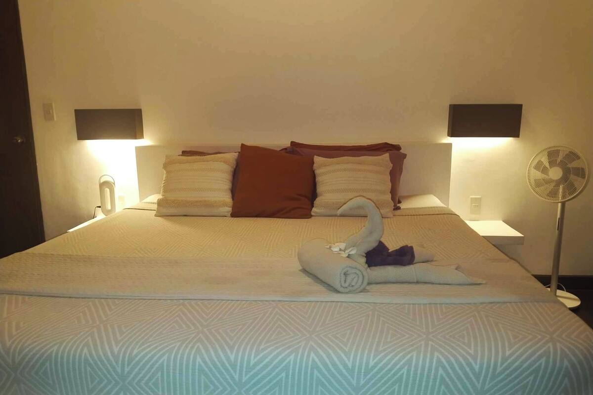 Cama bem arrumada com decoração de toalha em formato de cisne, duas luminárias de cabeceira e um ventilador de pé.