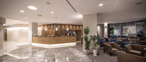 Um hall de hotel moderno com balcão de recepção, área de estar e uma grande planta em vaso.