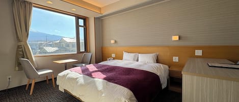 Quarto de hotel com cama, escrivaninha, cadeira e janela com vista para edifícios e montanhas.