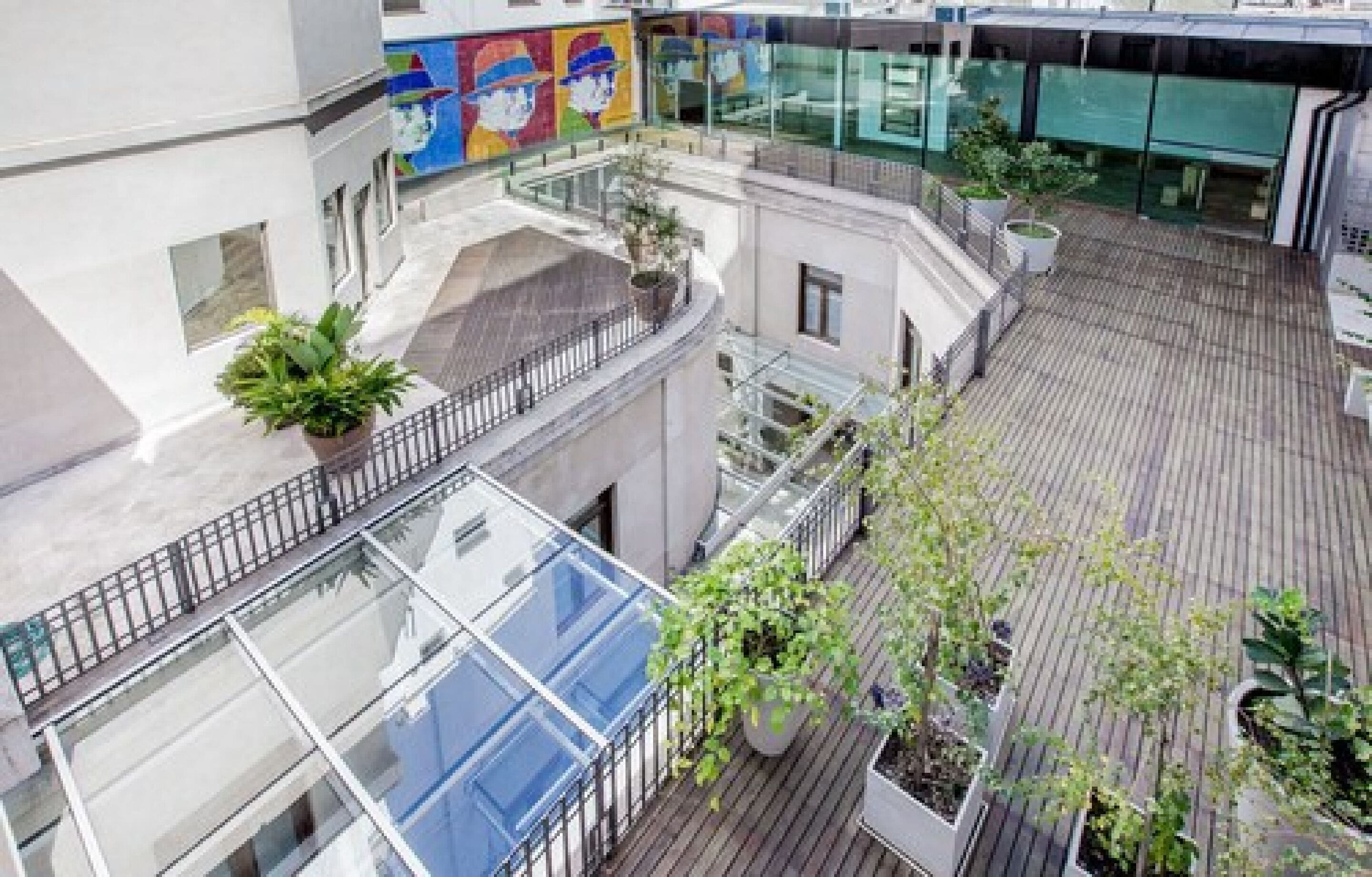 Terraço ao ar livre com vários níveis, guarda-corpos de vidro, plantas em vasos e um mural colorido na fachada do edifício.