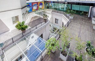 Terraço ao ar livre com vários níveis, guarda-corpos de vidro, plantas em vasos e um mural colorido na fachada do edifício.