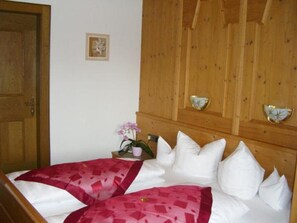 Quarto com cabeceira de madeira, cama com roupa de cama branca e rosa, mesa de cabeceira pequena com um vaso de flores e um quadro na parede.