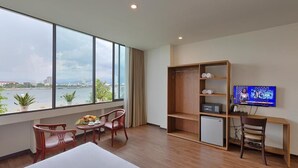 Quarto de hotel com uma janela grande, televisão, escrivaninha, cadeira, mesinha com flores e guarda-roupa.