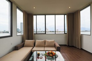 Quarto de hotel moderno com sofá, mesa de centro de vidro e vista para a paisagem urbana pelas amplas janelas.