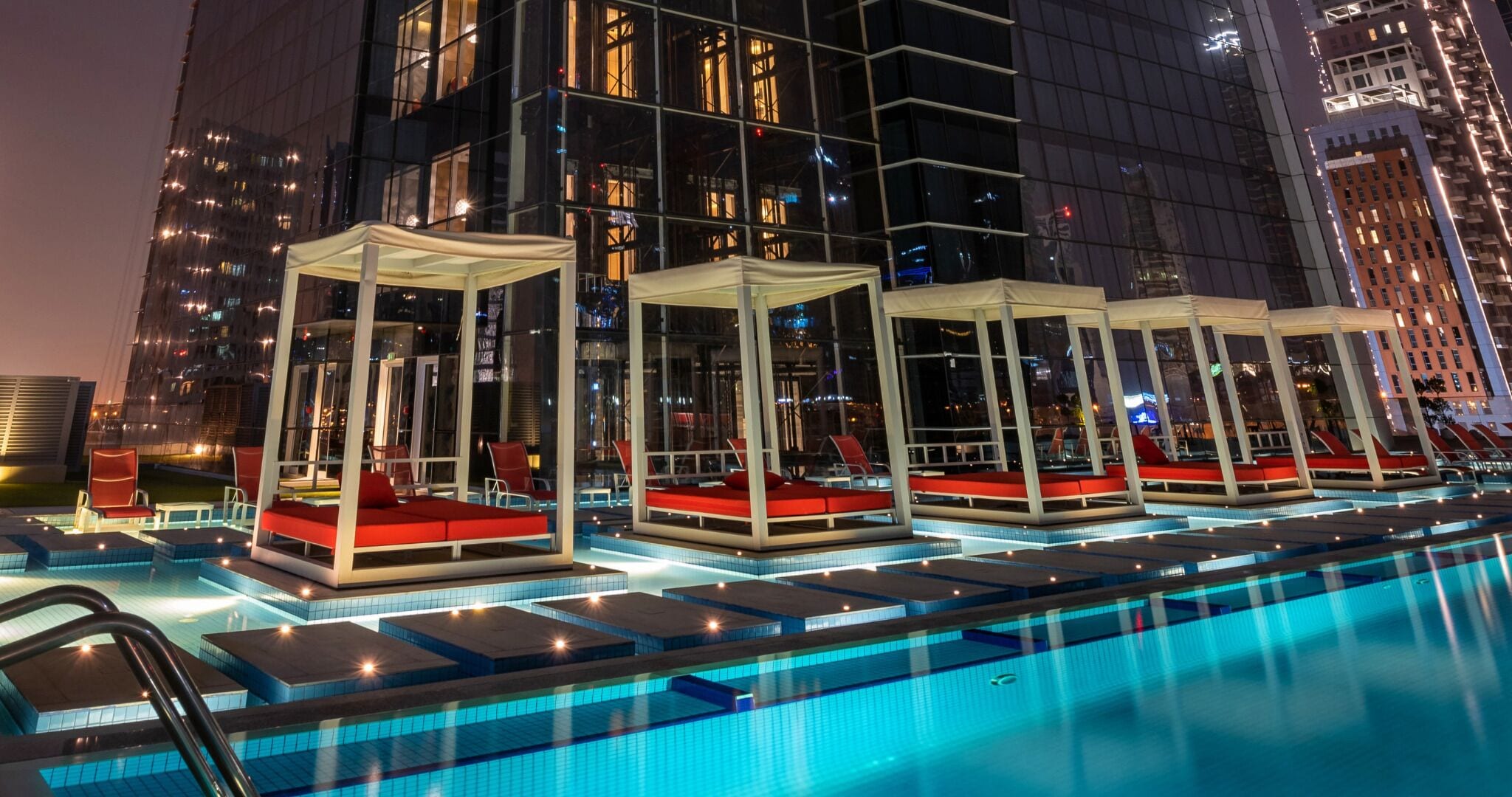 Piscina no terraço com várias espreguiçadeiras e áreas de estar cobertas, cercada por edifícios altos.
