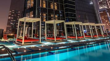 Piscina no terraço com várias espreguiçadeiras e áreas de estar cobertas, cercada por edifícios altos.