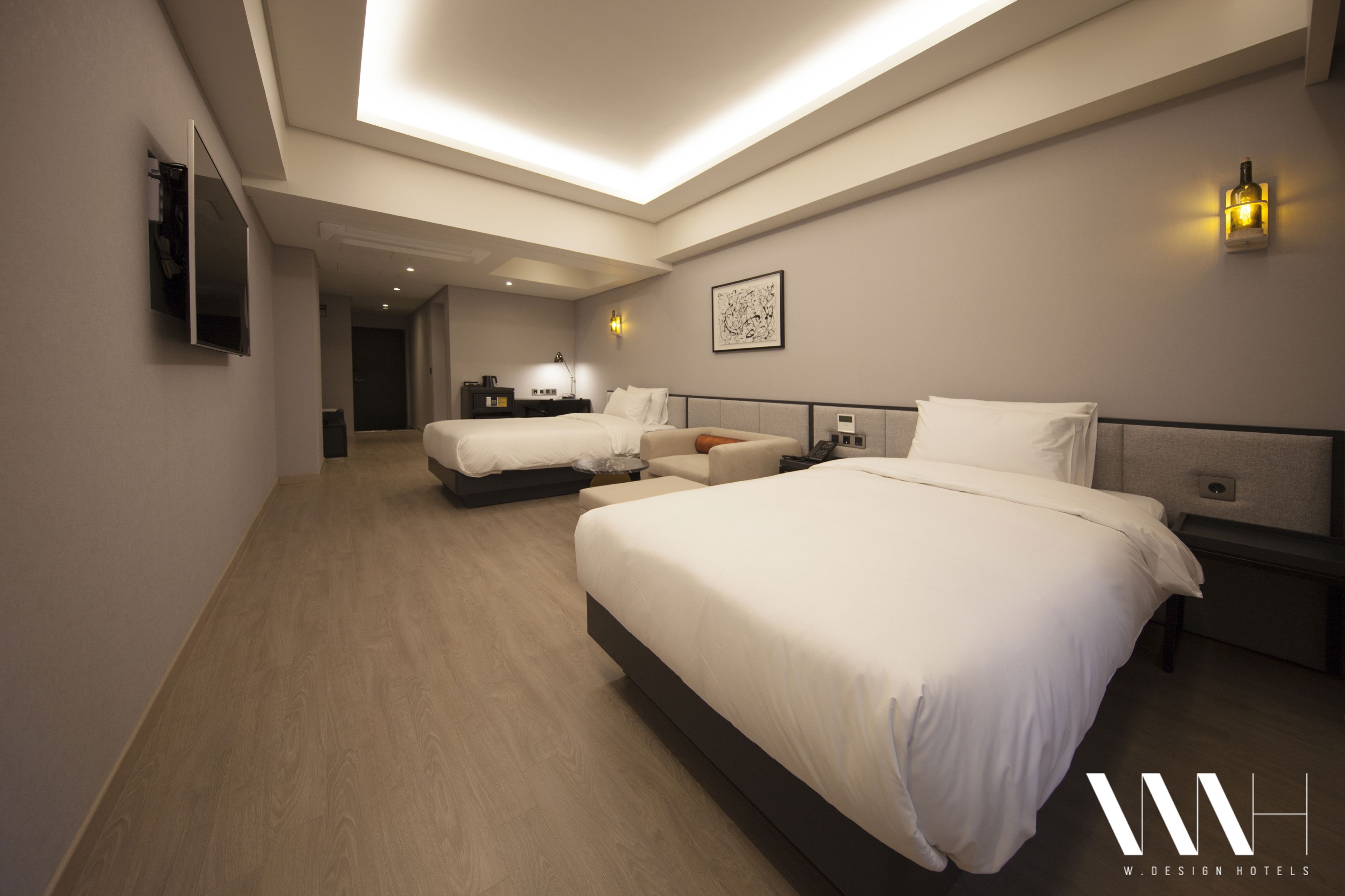 Quarto de hotel moderno com uma cama grande, uma cama menor e um sofá. Há uma escrivaninha com uma luminária, uma TV fixada na parede e um quadro emoldurado.