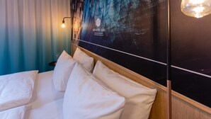Quarto de hotel com cama, almofadas brancas, uma obra de arte na parede e um arandelador.