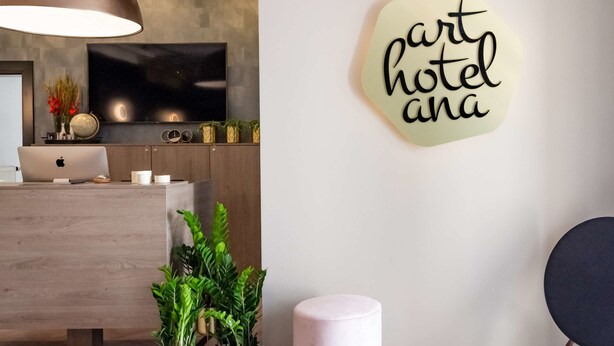 Uma área de recepção de hotel moderna com uma televisão de tela plana, um laptop e objetos decorativos sobre um balcão de madeira. Há uma placa fixada na parede com a inscrição "art hotel ana".