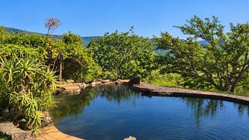 Área de piscina ao ar livre tranquila, com céu azul límpido, cercada por vegetação exuberante e montanhas ao fundo.