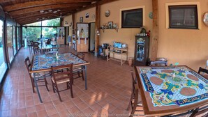 Um alpendre espaçoso com vigas de madeira, soalho em mosaico e mesas com toalhas de mesa coloridas.