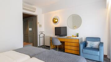 Quarto de hotel com duas camas, uma escrivaninha com computador, uma cadeira, uma poltrona azul, um espelho redondo e um radiador.
