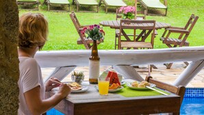 Uma sala de jantar com uma mesa posta para dois, um vaso de flores e vista para a piscina.