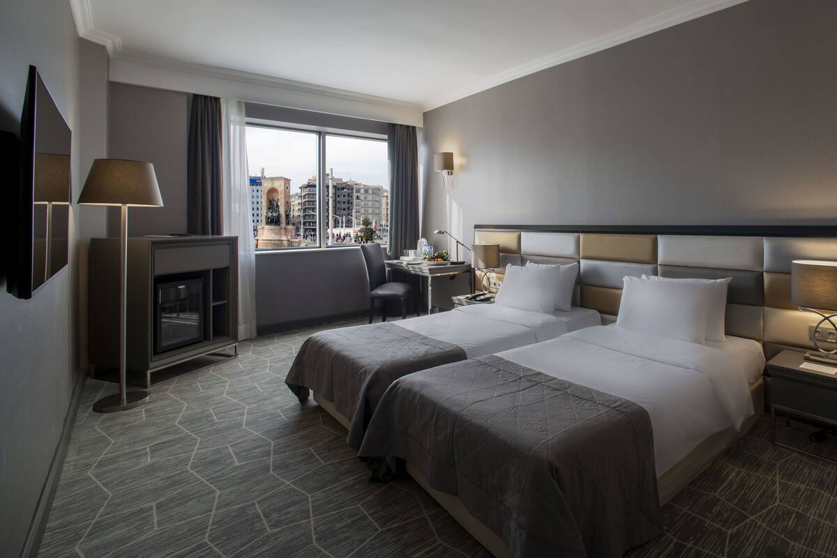 Quarto de hotel com duas camas, uma mesa, uma cadeira, televisão e uma janela grande com vista para a cidade.