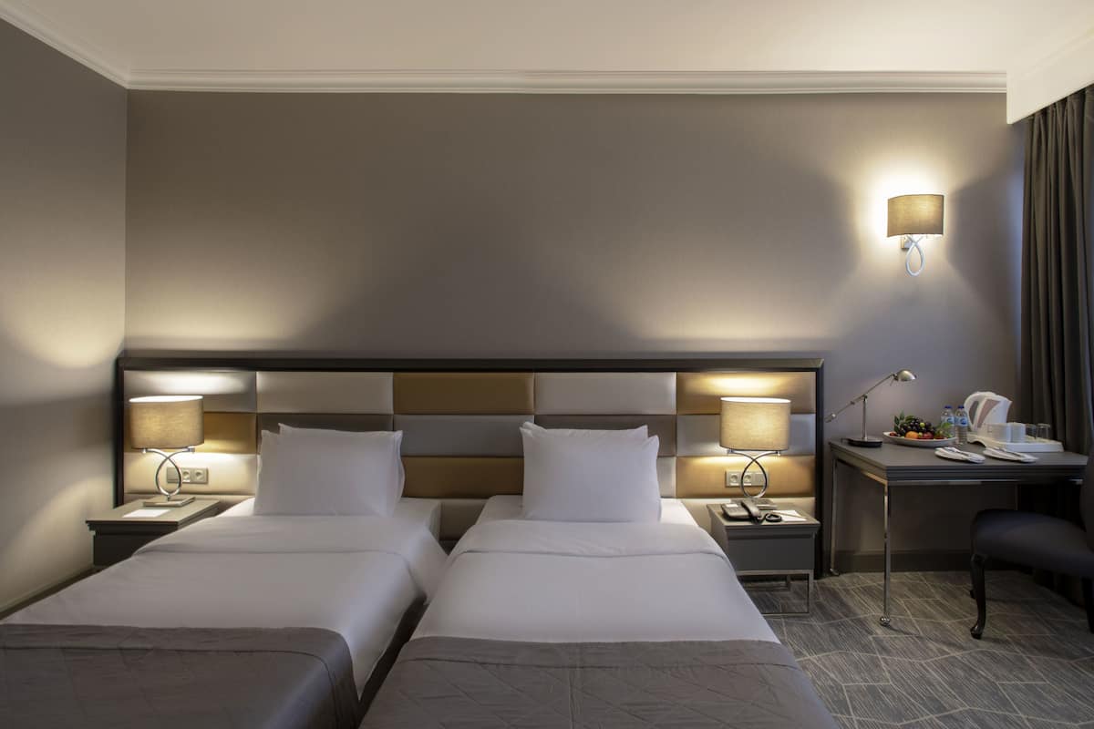 Quarto de hotel com duas camas, uma escrivaninha com abajur, uma cadeira e uma mesinha de cabeceira com telefone e luminária.