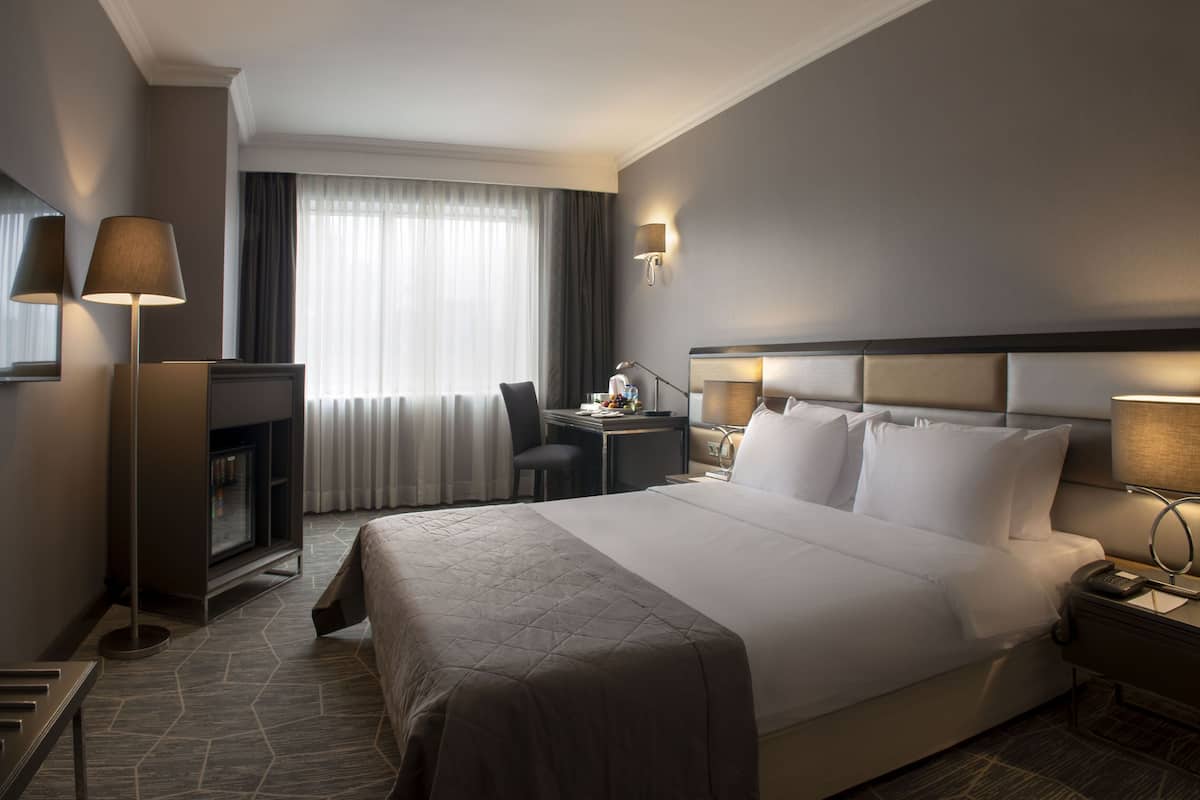 Quarto de hotel com uma cama grande, uma escrivaninha com cadeira, televisão e luminária.