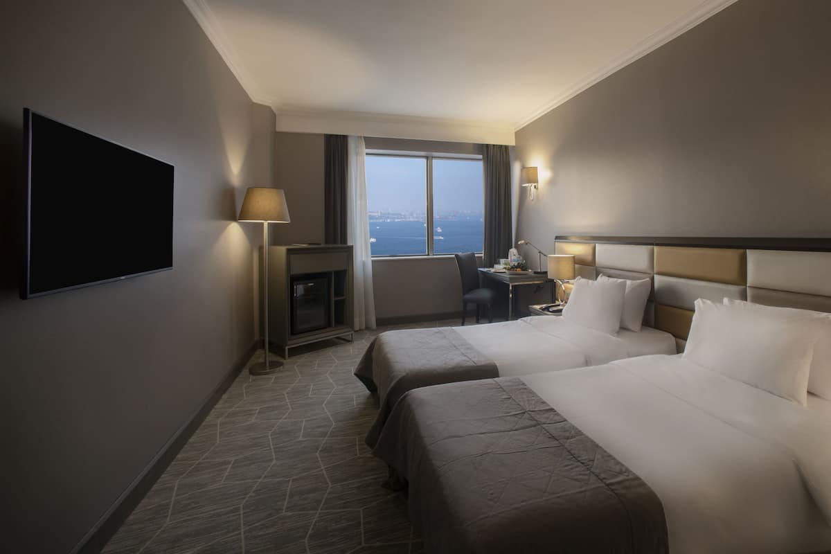 Quarto de hotel com duas camas, televisão, escrivaninha, cadeira, luminária e vista da cidade através de uma janela grande.