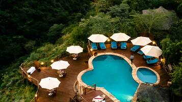 Piscina ao ar livre rodeada por espreguiçadeiras e guarda-sóis, construída sobre um deck de madeira com floresta ao fundo.