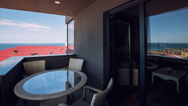 Uma varanda com mesa de vidro e cadeiras de vime, com vista para a paisagem urbana costeira.