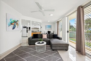 Uma sala de estar moderna com sofá modular, mesa de centro redonda e ventilador de teto. Cozinha visível ao fundo, e varanda com vista.