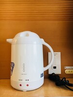 Uma chaleira elétrica branca com tampa, colocada sobre uma superfície de madeira ao lado de uma tomada.