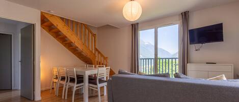 Uma sala de estar moderna com escada de madeira, área de jantar, sofá cinza e TV fixada na parede.