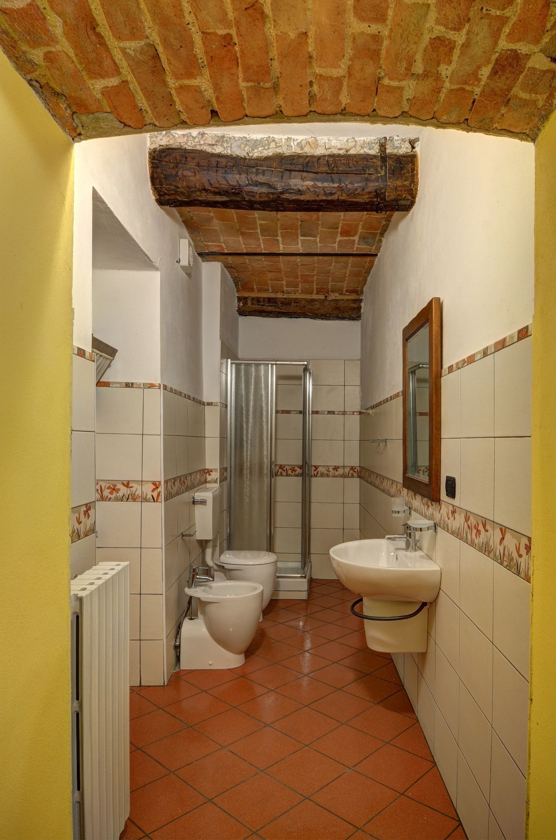 Casa de banho com chuveiro, sanita e lavatório, com paredes em azulejo e teto de tijolo.