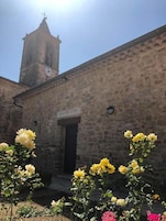 Um edifício de pedra com uma torre de relógio, uma placa com a inscrição "GALERIE D'ART", e um jardim com flores amarelas.