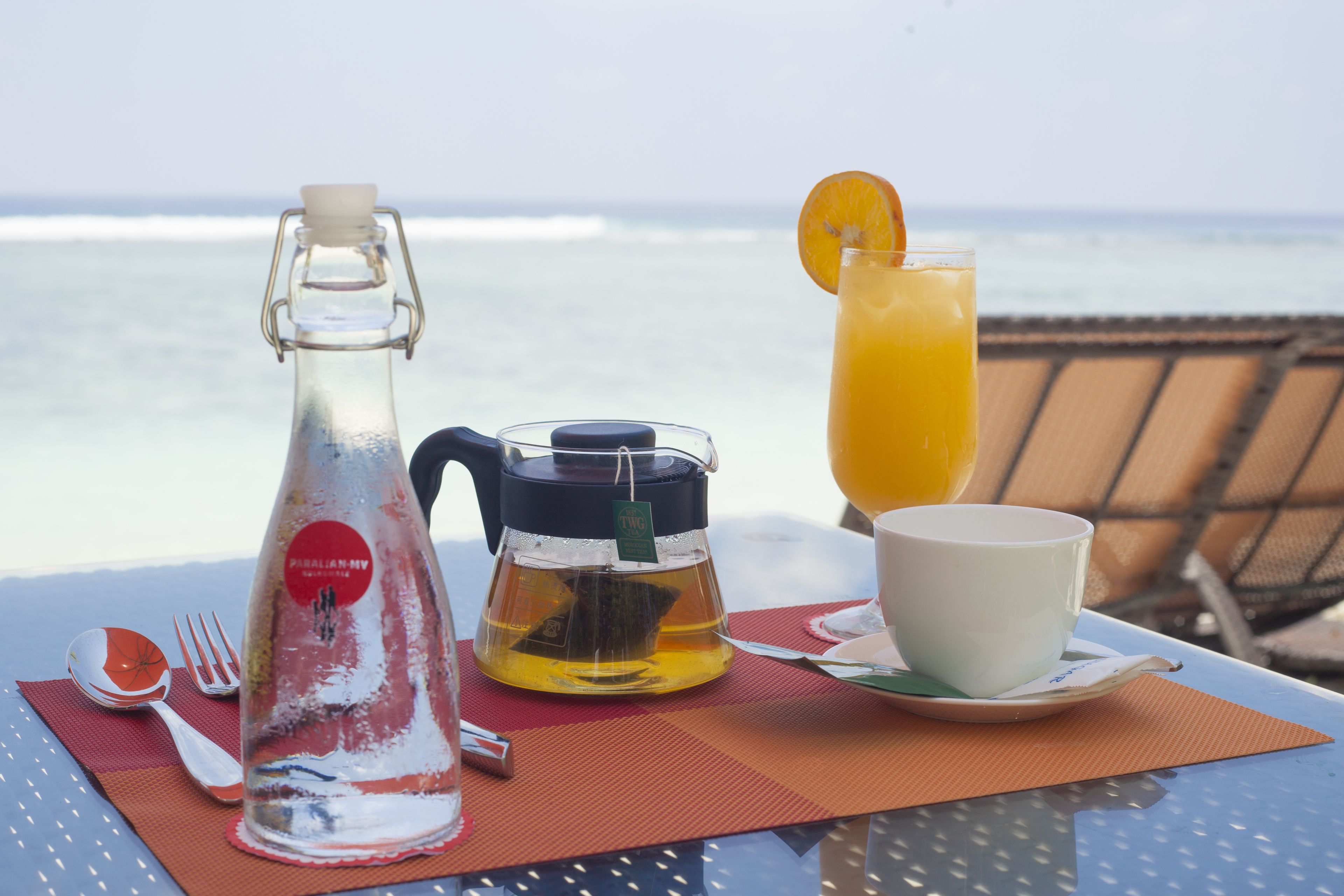 Uma mesa com uma garrafa, uma chaleira e um copo de sumo de laranja, numa praia com vista para o oceano.