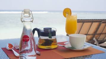 Uma mesa com uma garrafa, uma chaleira e um copo de sumo de laranja, numa praia com vista para o oceano.