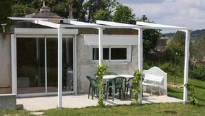 Um alpendre coberto com mesa, cadeiras e um banco, além de uma estrutura de pérgola.