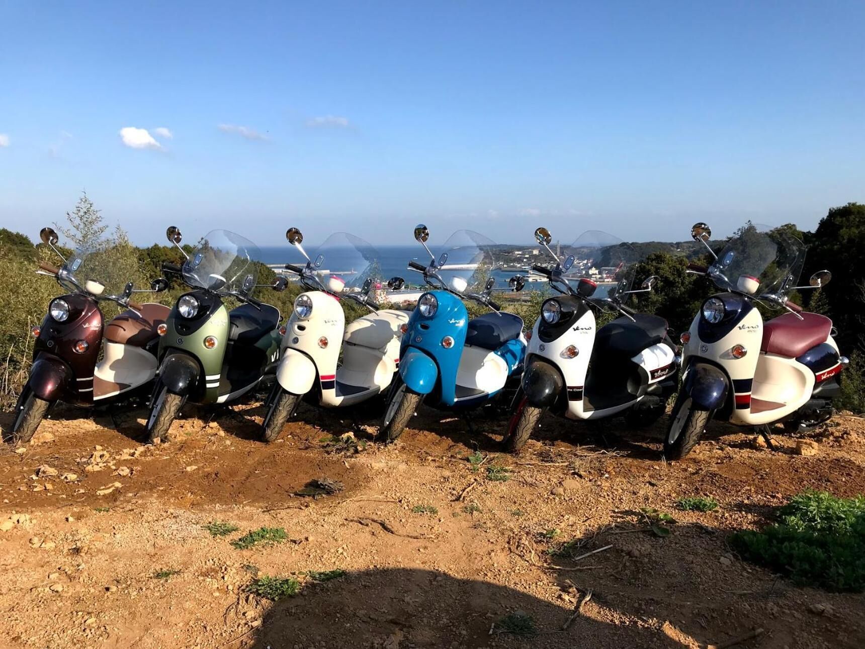 Fileira de scooters estacionadas numa trilha de terra com vista panorâmica de uma cidade costeira ao fundo.