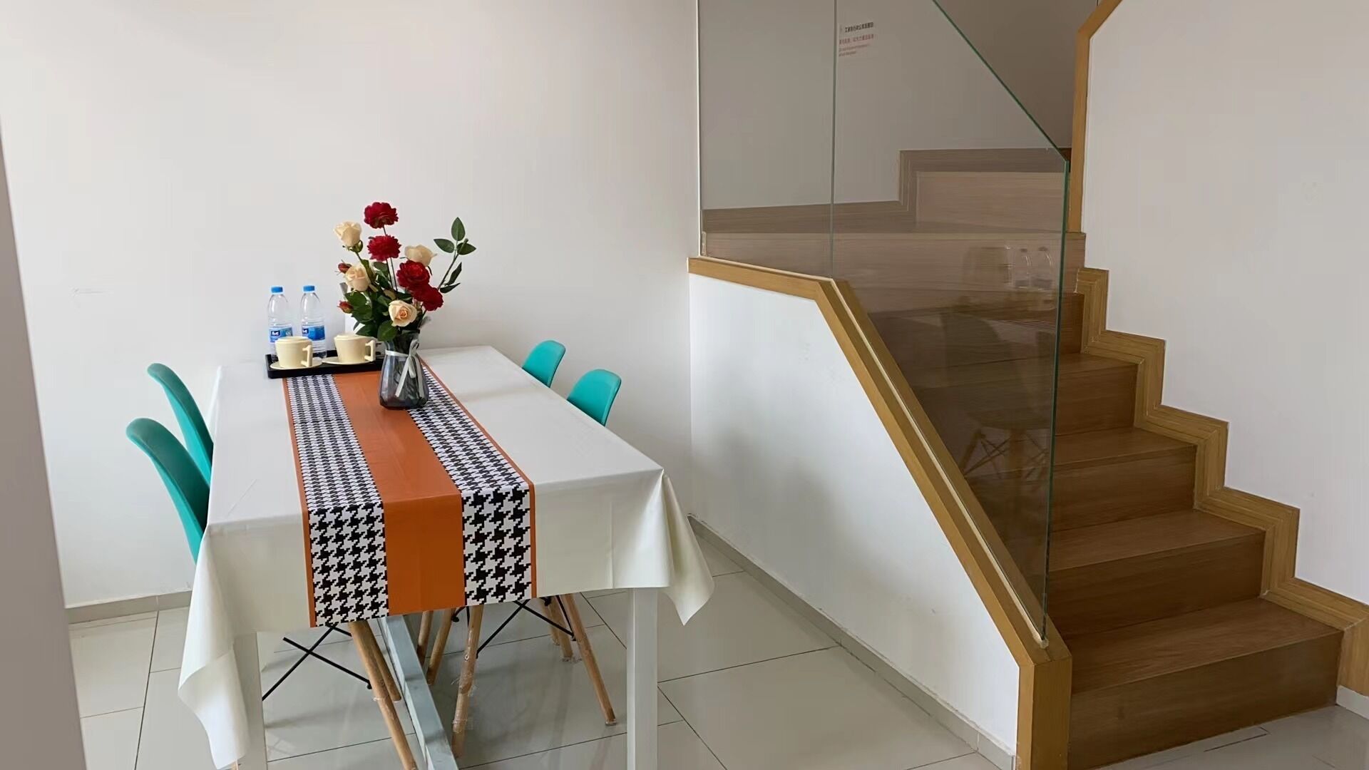 Uma área de refeições com uma mesa posta para quatro pessoas, um vaso de flores e uma escadaria com guarda-corpos de vidro.