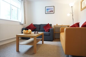 Uma sala de estar com um sofá azul, uma mesa de centro de madeira com frutas e um muffin, uma poltrona amarela e um quadro na parede.
