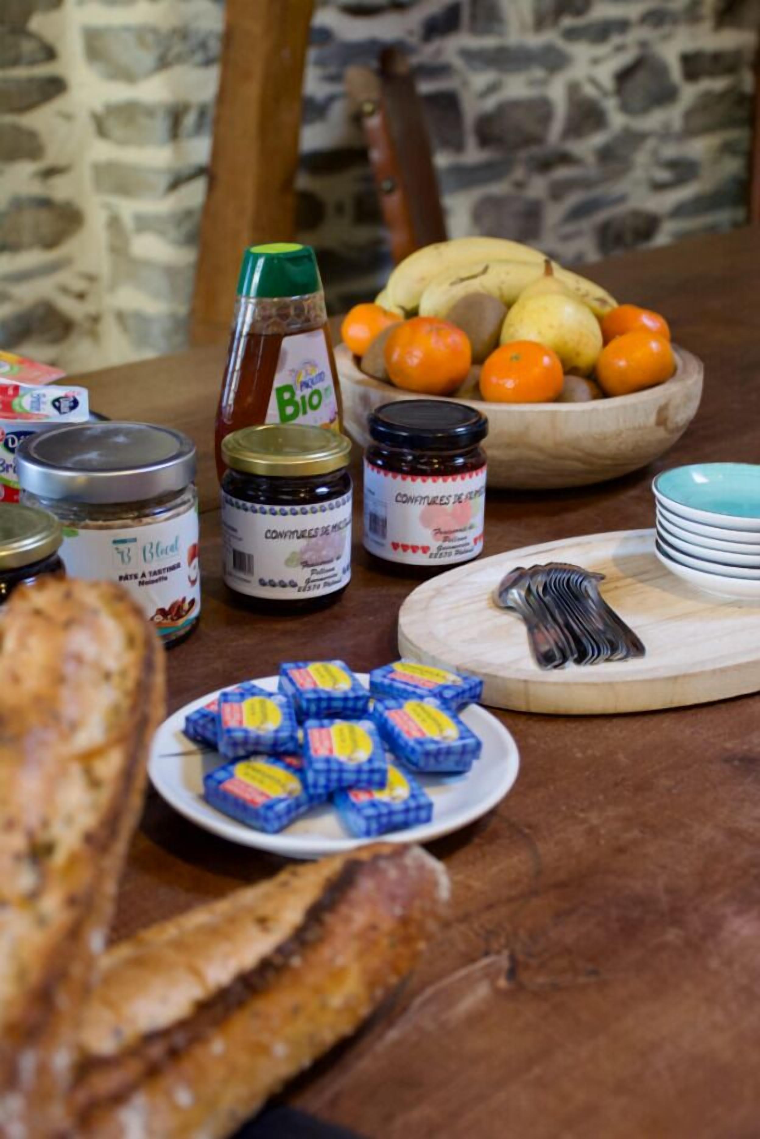 Uma mesa com diversos alimentos, incluindo uma tigela de laranjas e bananas, potes de compotas e um prato de lanches embalados em azul e amarelo.