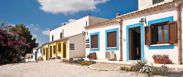 Fileira de edifícios brancos com molduras de janelas azuis e portas amarelas, situada em um pátio ensolarado com caminhos de paralelepípedos e plantas em vasos.