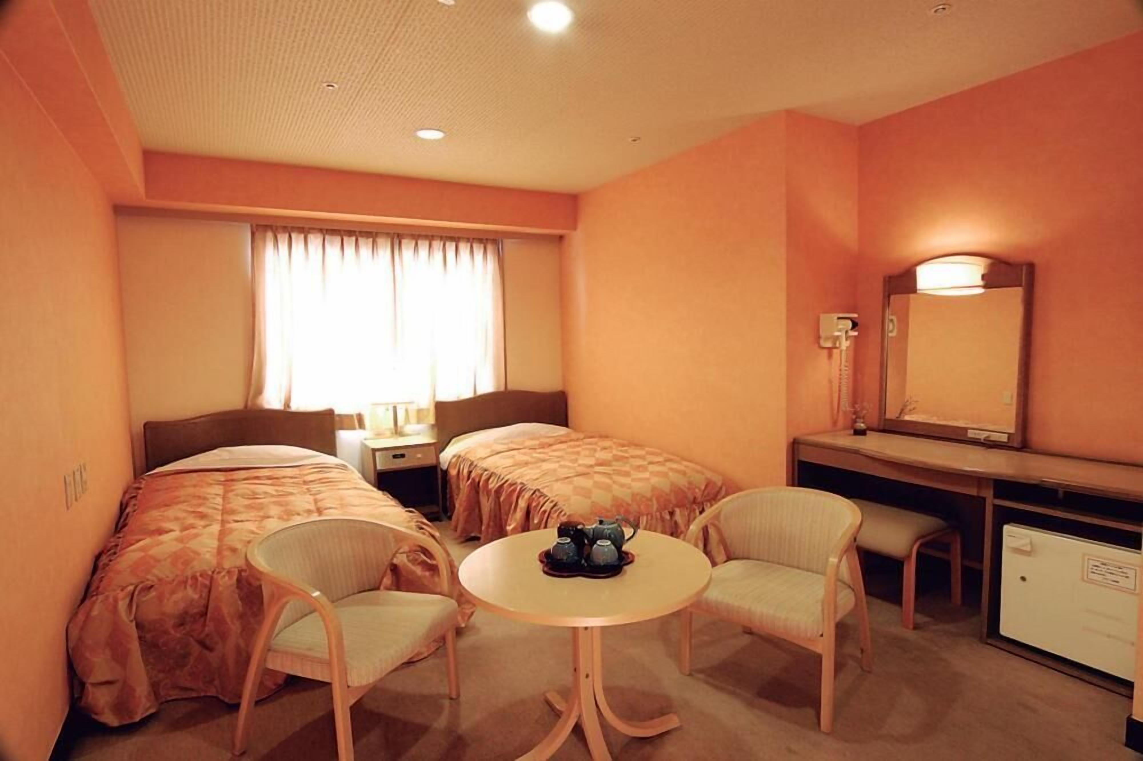Quarto de hotel com duas camas de solteiro, uma mesa redonda pequena com um bule, duas cadeiras, uma escrivaninha com um computador, um espelho e uma janela com cortinas.
