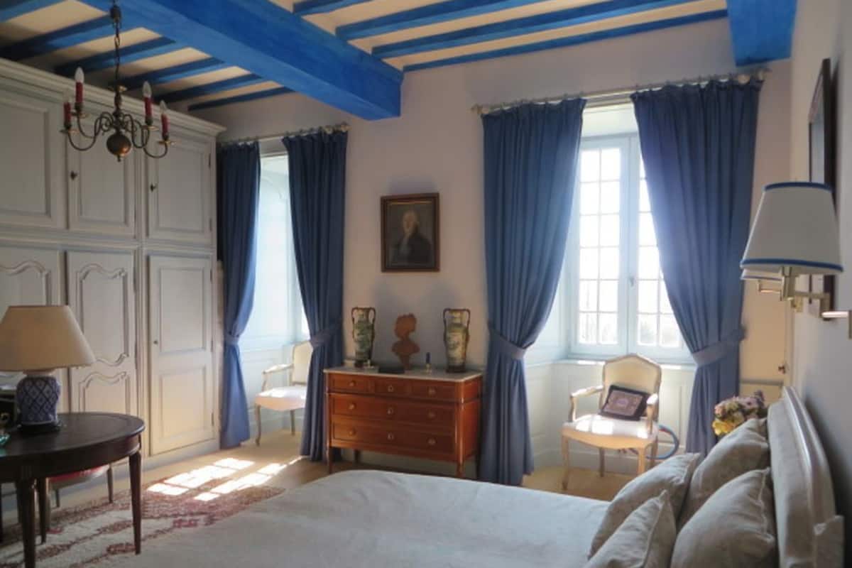 Quarto com cama, cômoda e duas cadeiras. Há um lustre, uma janela com cortinas e um abajur sobre uma mesa.