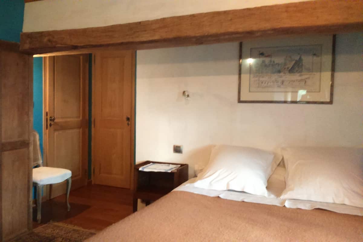 Quarto com teto de madeira, cama com roupa de cama branca, mesa de cabeceira, cadeira e porta que dá para outro cômodo.