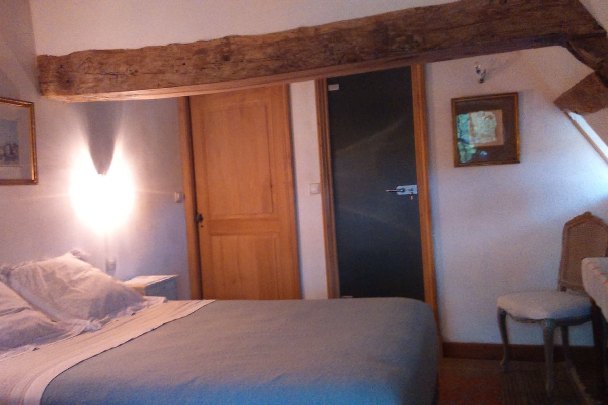 Um quarto com cama, porta de madeira, uma cadeira e duas imagens emolduradas na parede.