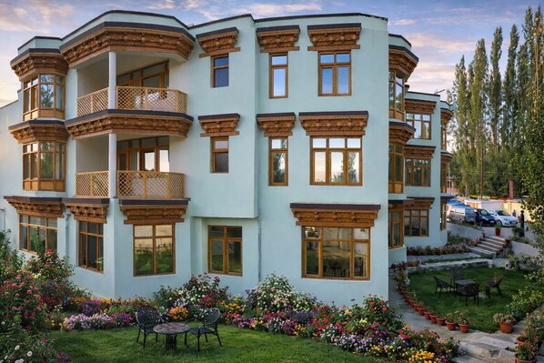 Um edifício de vários andares com fachada azul clara, várias varandas com grades de madeira e um jardim bem cuidado, com flores coloridas e mobiliário de exterior.