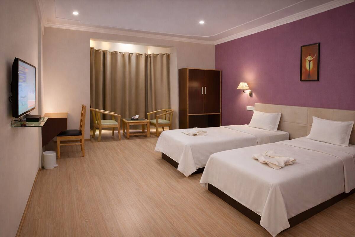 Quarto de hotel com duas camas, uma mesa com computador, uma cadeira, guarda-roupa e um quadro na parede.