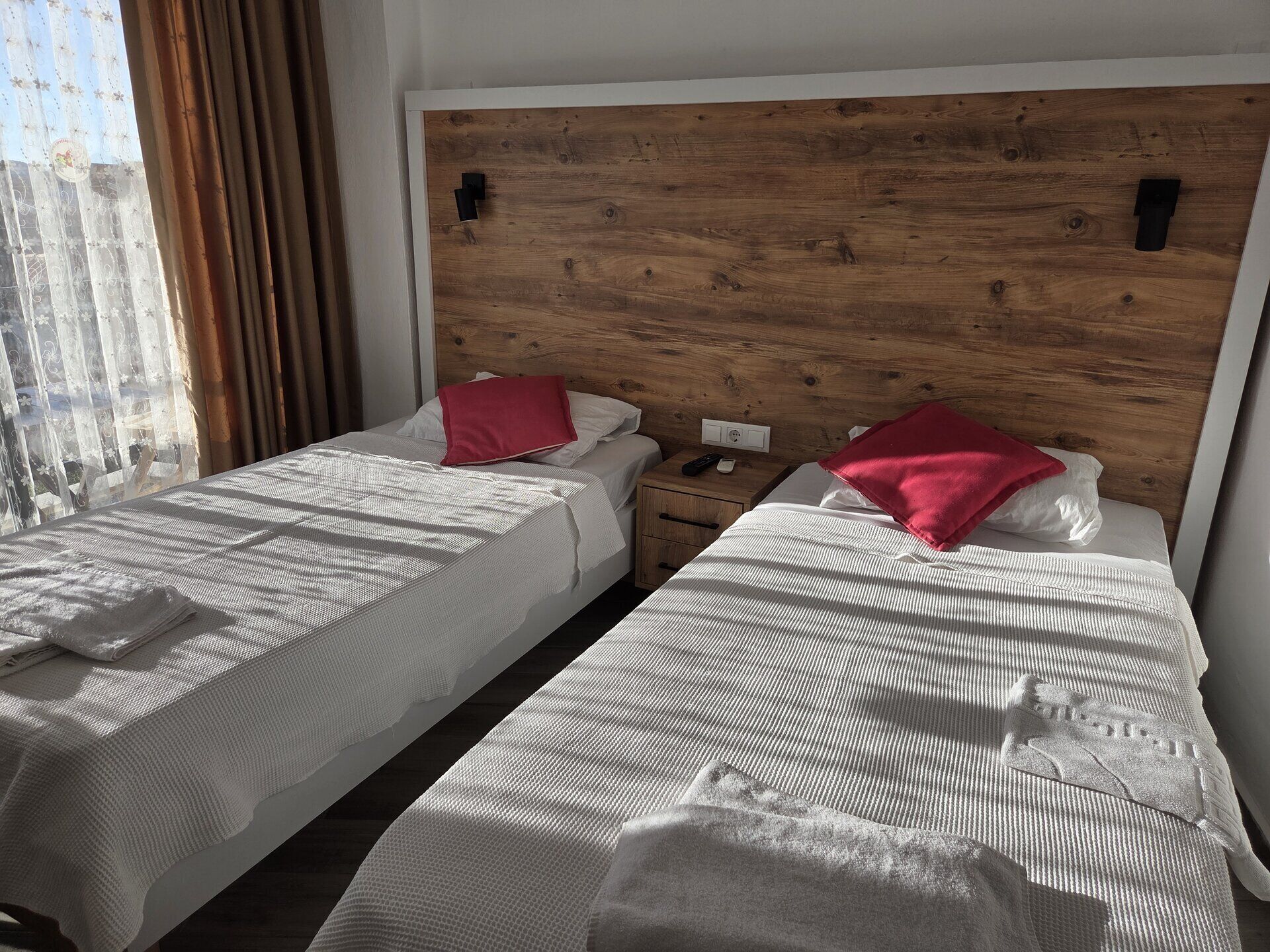 Quarto de hotel com duas camas de solteiro, cabeceira de madeira, mesa de cabeceira com telefone e janela com cortinas.