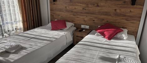 Quarto de hotel com duas camas de solteiro, cabeceira de madeira, mesa de cabeceira com telefone e janela com cortinas.