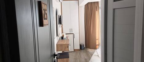 Quarto de hotel com cama, escrivaninha de madeira, espelho, guarda-roupa e janela com cortinas.