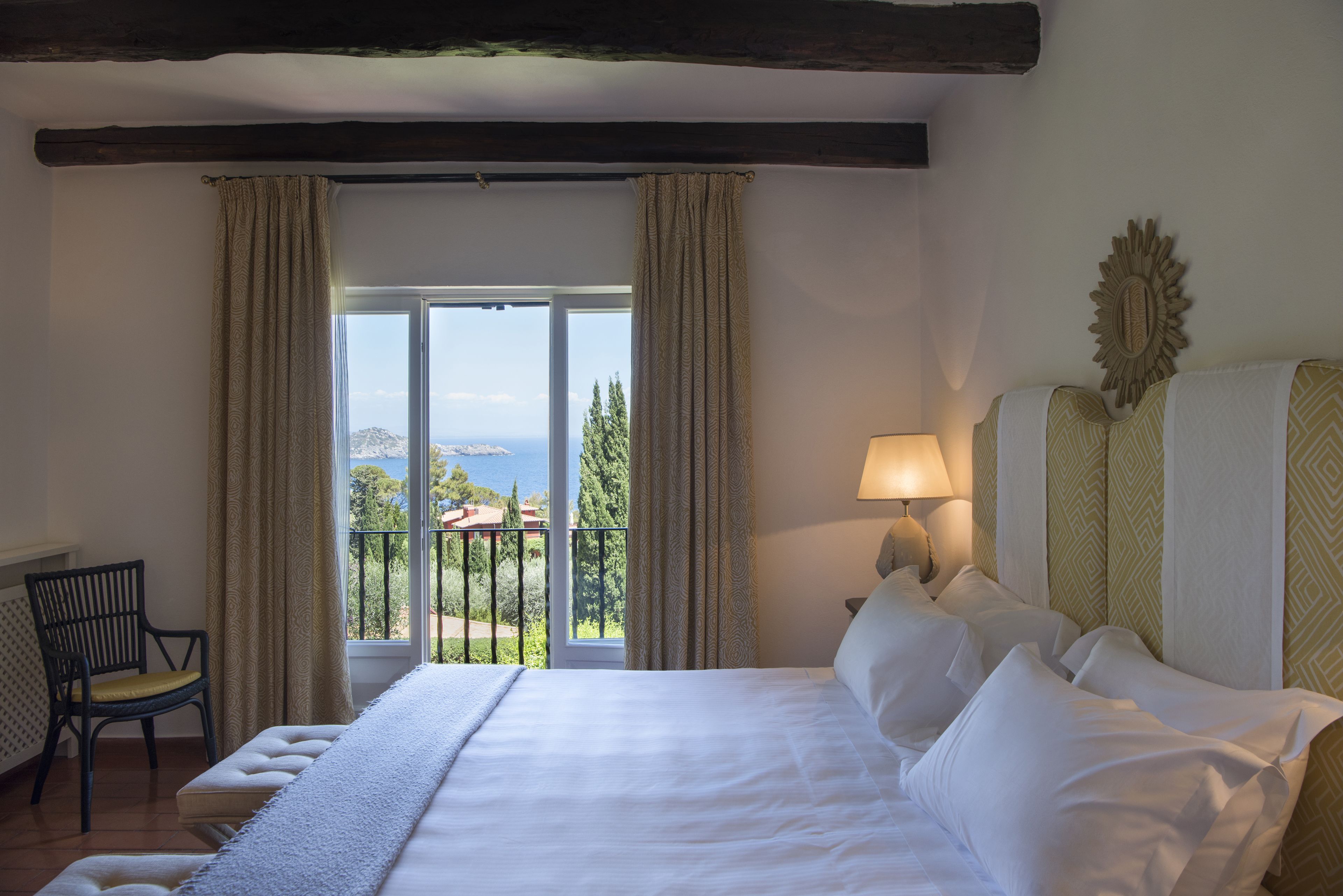 Quarto de hotel com cama, uma cadeira, abajur e vista para o mar através de uma janela grande.