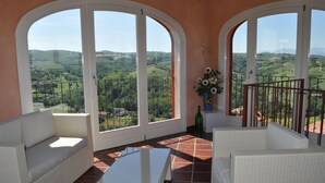 Uma varanda com móveis de vime branco, uma mesa de vidro e vista para colinas verdes.