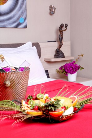 Quarto de hotel bem arrumado, com uma cama, um painel decorativo na parede, uma pequena estante com uma estátua, um vaso com flores e uma cesta de frutas sobre a cama.