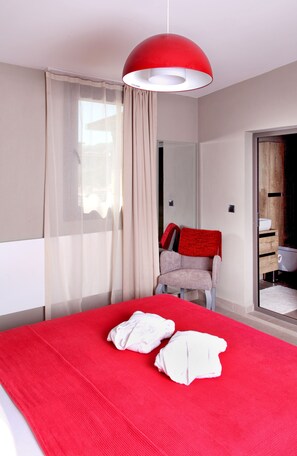 Quarto de hotel com cama, luminária pendente vermelha e branca, uma cadeira, manta vermelha e banheiro visível por uma porta aberta.
