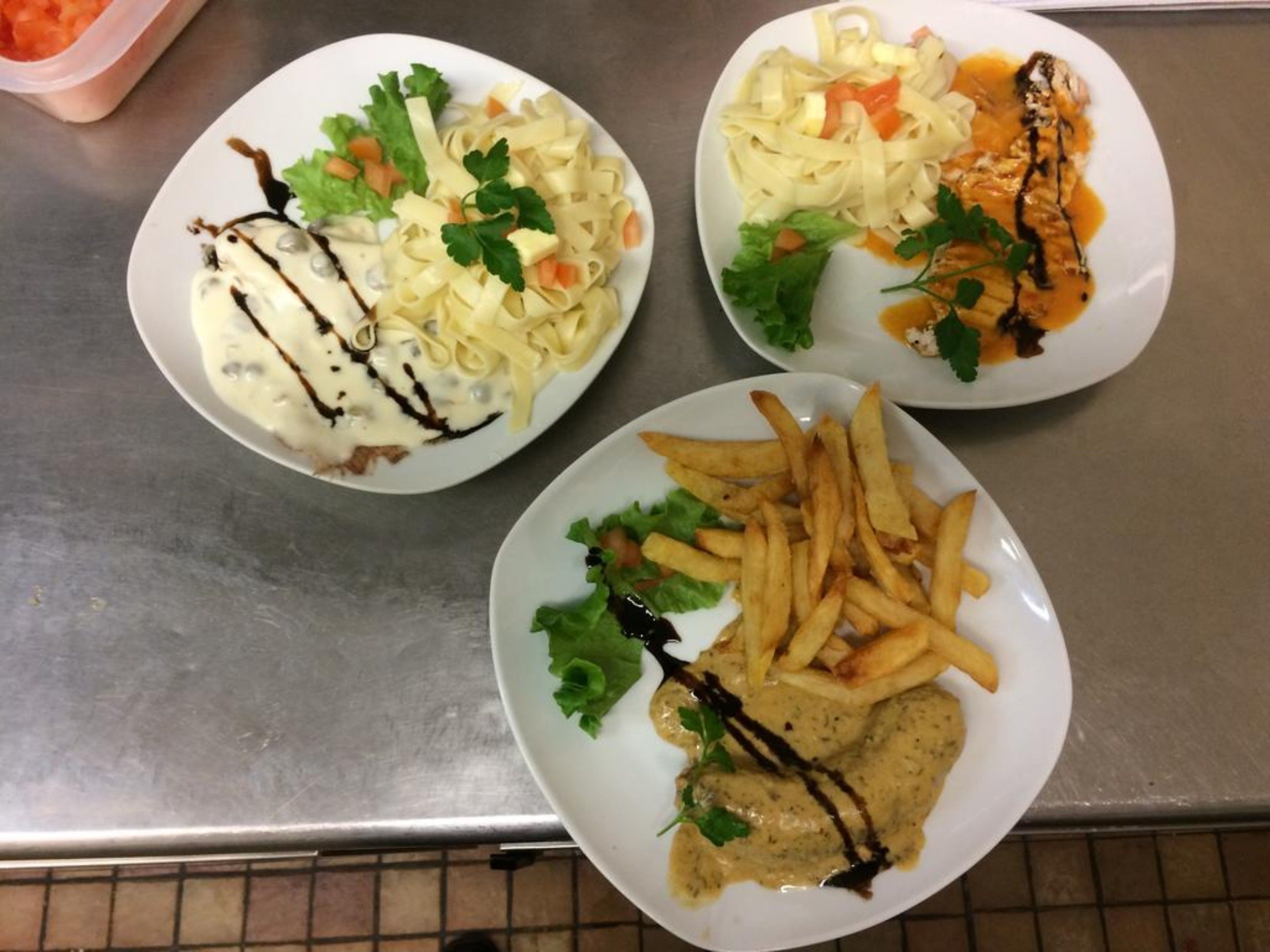 Três pratos de comida com diferentes pratos, incluindo massa, macarrão e batatas fritas, sobre uma superfície metálica.